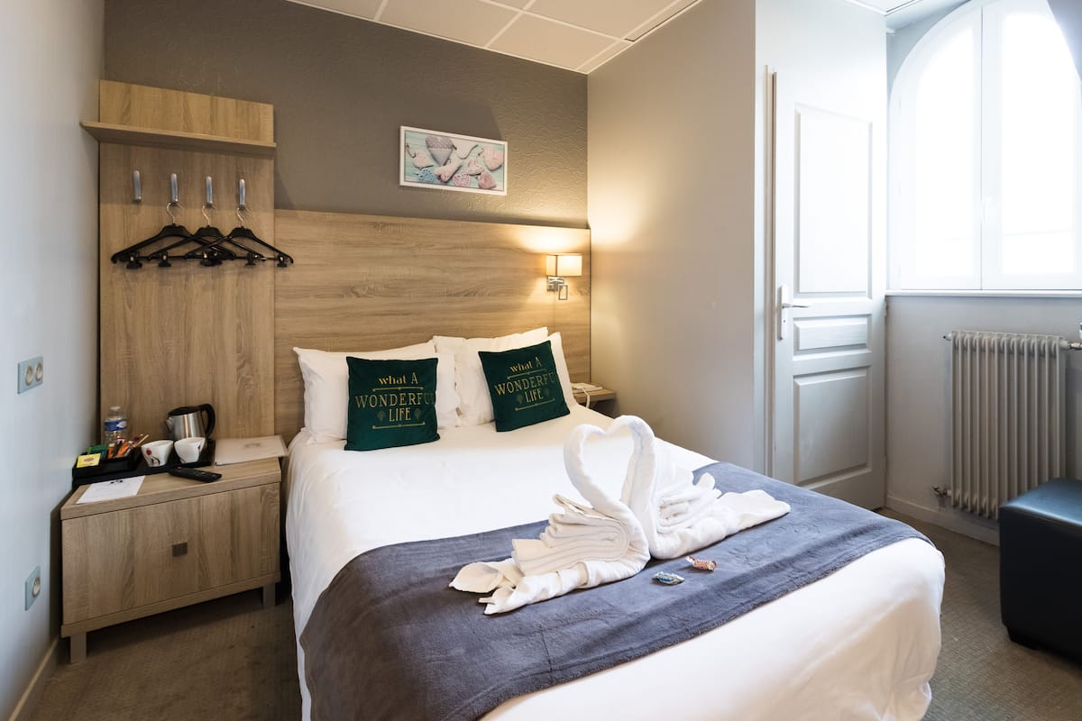 Quarto de hotel com cama arrumada, guarda-roupa de madeira, cabideiro na parede, mesa de cabeceira com chaleira e janela com vista.