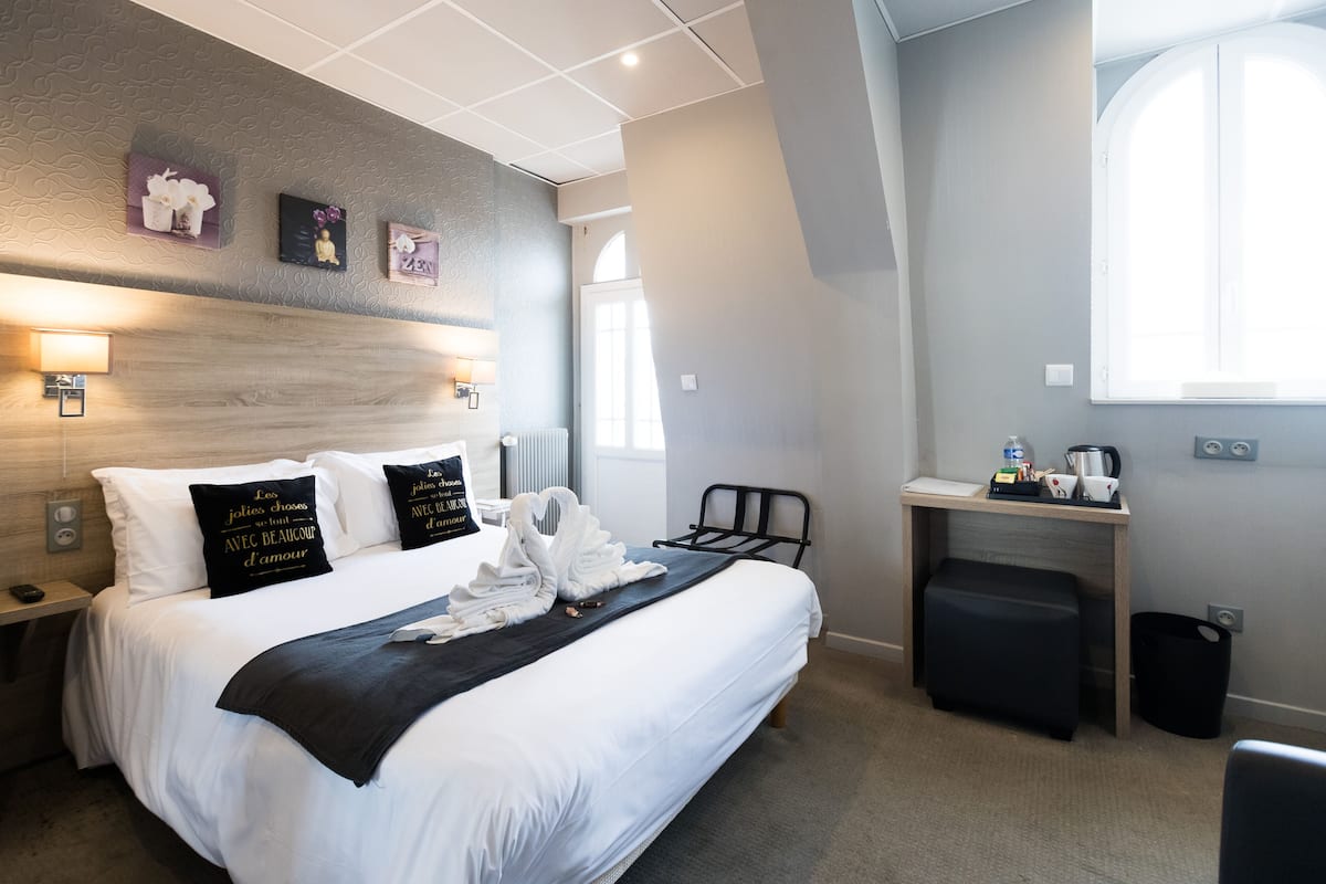 Quarto de hotel com cama bem arrumada, uma escrivaninha com cafeteira e uma lixeira.