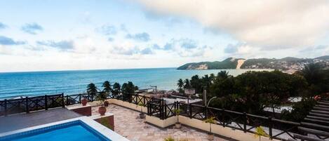 Piscina no terraço com vista para o oceano e uma cidade ao longe.