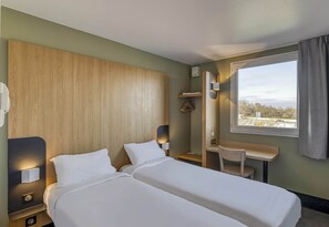 Quarto de hotel com duas camas, uma escrivaninha, uma cadeira, janela com vista e cabeceira de madeira.