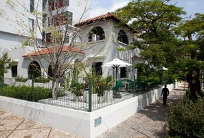 Um edifício branco com janelas em arco e telhado telhado, cercado por um muro branco e vegetação.