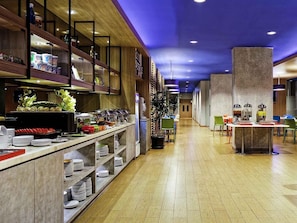 Um hall de hotel moderno com um balcão de recepção extenso, uma área de buffet com diversas opções de comida e um espaço de estar com mesas e cadeiras.