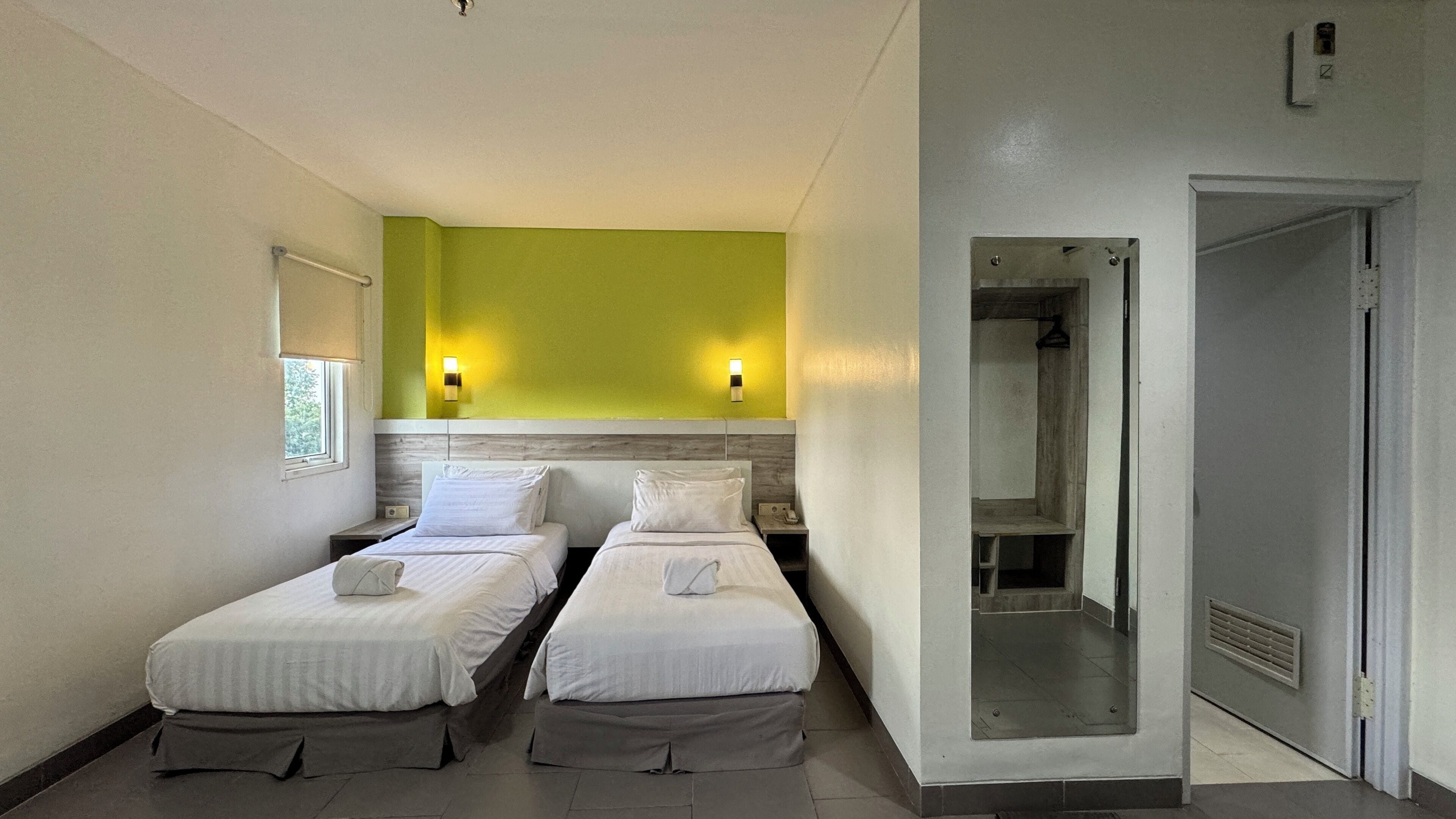 Quarto de hotel com duas camas de solteiro, um espelho, mesa de cabeceira com abajur e uma parede verde decorativa.