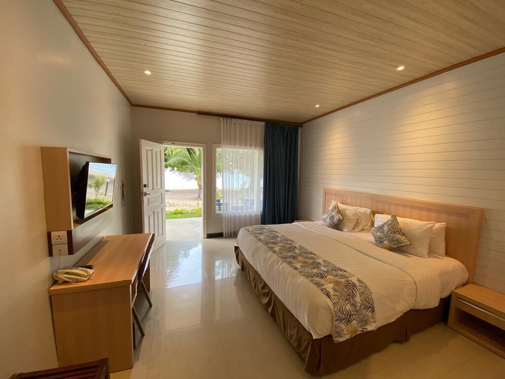 Quarto de hotel com teto de madeira, cama com colcha florida, televisão em suporte de madeira e vista para um jardim através de uma porta.