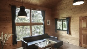 Interior acolhedor de cabana, com piso de madeira, sofá, mesa de centro de vidro e janelas grandes com cortinas.