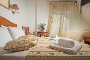 Quarto de hotel com cama, mesa de cabeceira, luminária, planta em vaso e janela com cortinas.