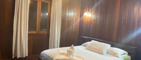 Uma cama bem arrumada com roupa de cama e toalha brancos, paredes de madeira, uma janela com cortinas e um lustre.