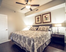 Quarto com cama, duas luminárias de cabeceira, um ventilador de teto e dois quadros botânicos na parede.