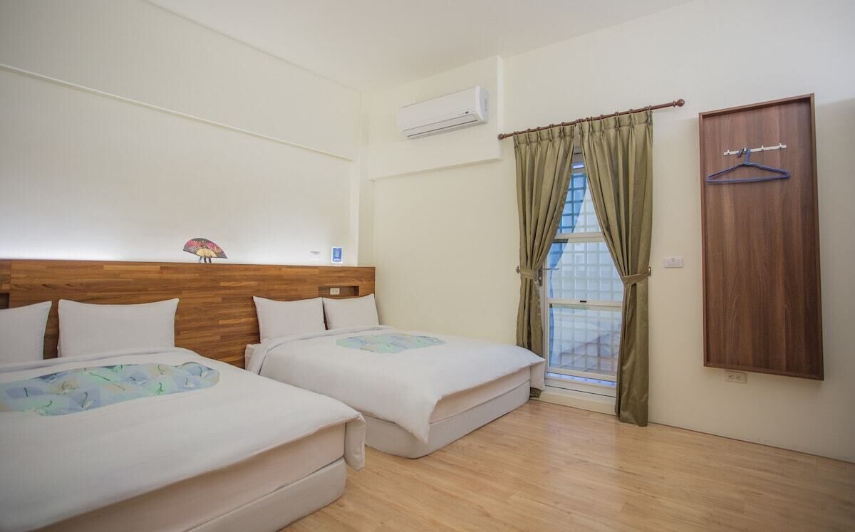 Quarto de hotel com duas camas, cabeceira de madeira, ar condicionado, janela com cortinas e guarda-roupa de madeira com cabide.