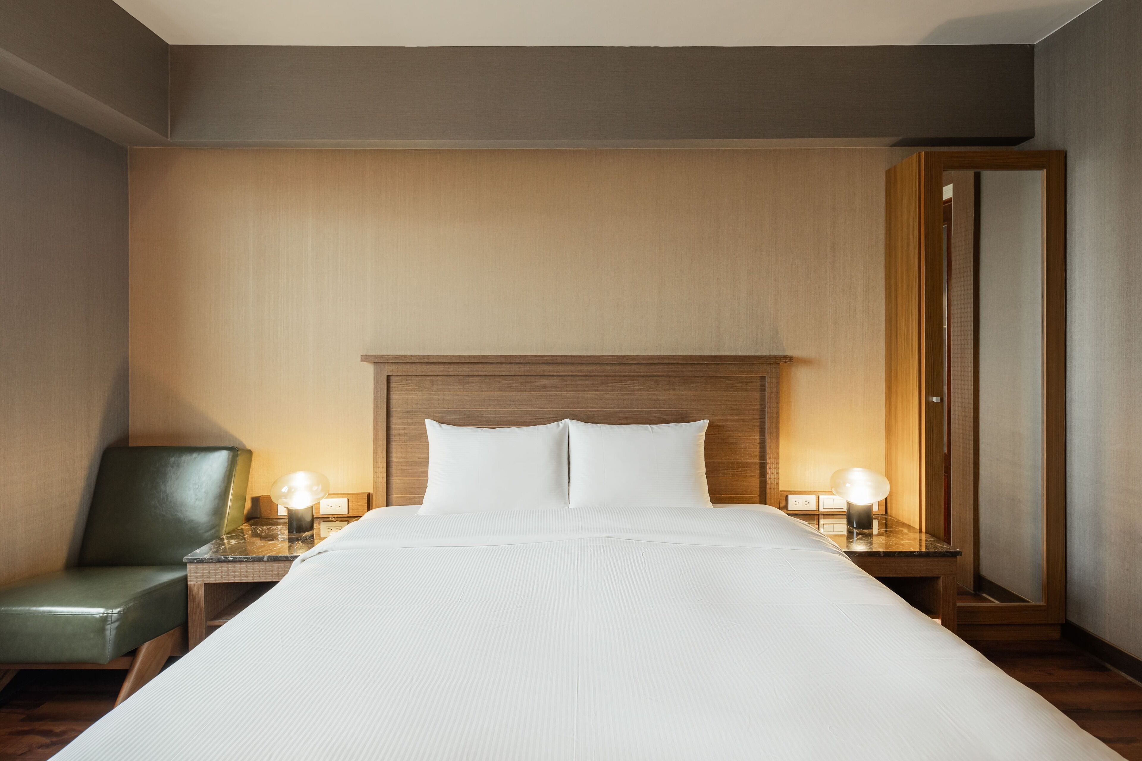 Quarto de hotel com uma cama grande, duas mesas de cabeceira com abajures, uma cadeira verde e uma cabeceira de madeira.