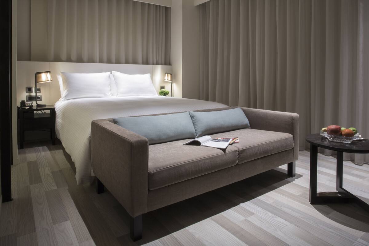 Quarto de hotel moderno com uma cama grande, um sofá, uma pequena mesa com frutas e uma mesa de cabeceira com luminárias.