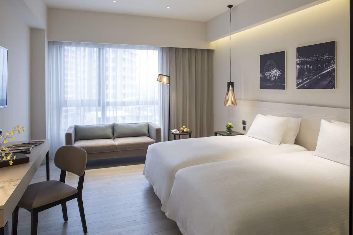 Quarto de hotel moderno com cama grande, sofá, escrivaninha com cadeira e vista para a cidade através de amplas janelas.
