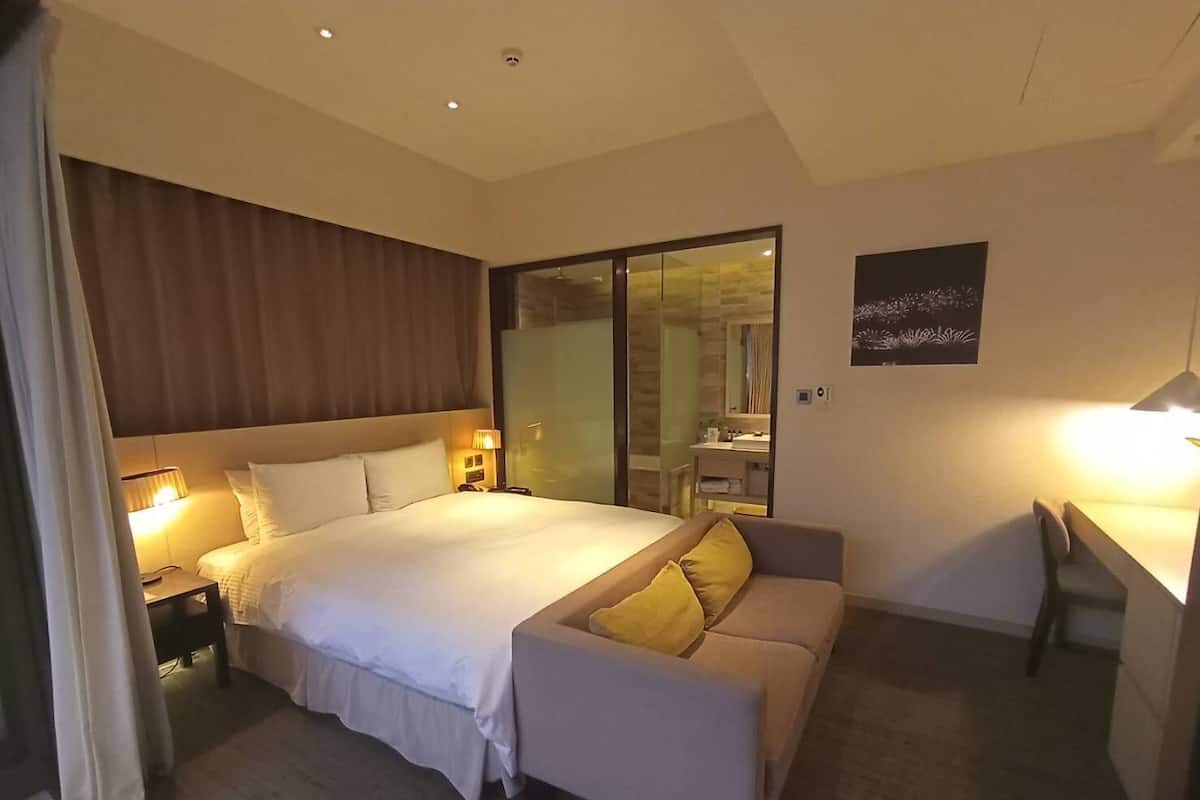 Quarto de hotel moderno com uma cama grande, um sofá, uma escrivaninha e um banheiro visível através de uma porta de vidro.