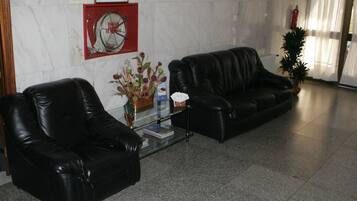 Um hall com dois sofás de couro preto, uma mesa de centro de vidro, um vaso com flores secas e um extintor fixado na parede.