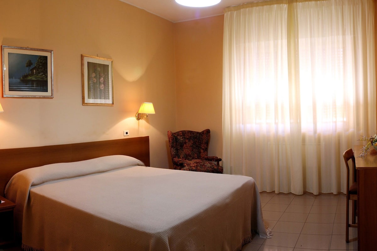Quarto de hotel com cama, duas luminárias na parede, uma cadeira, escrivaninha com uma tigela de frutas e televisão fixada na parede.