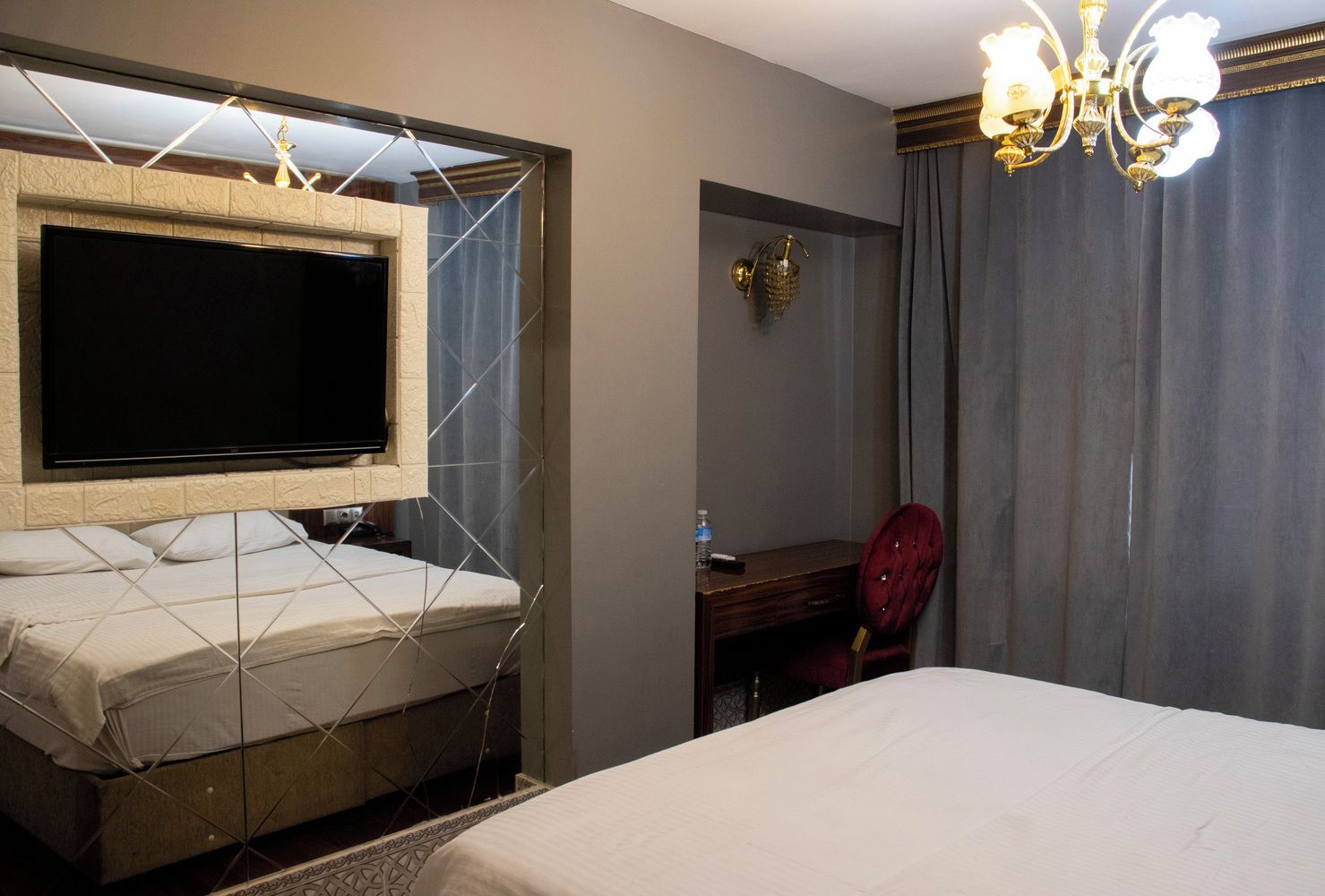Quarto de hotel com uma cama grande, televisão fixada na parede, escrivaninha com cadeira e um candelabro.