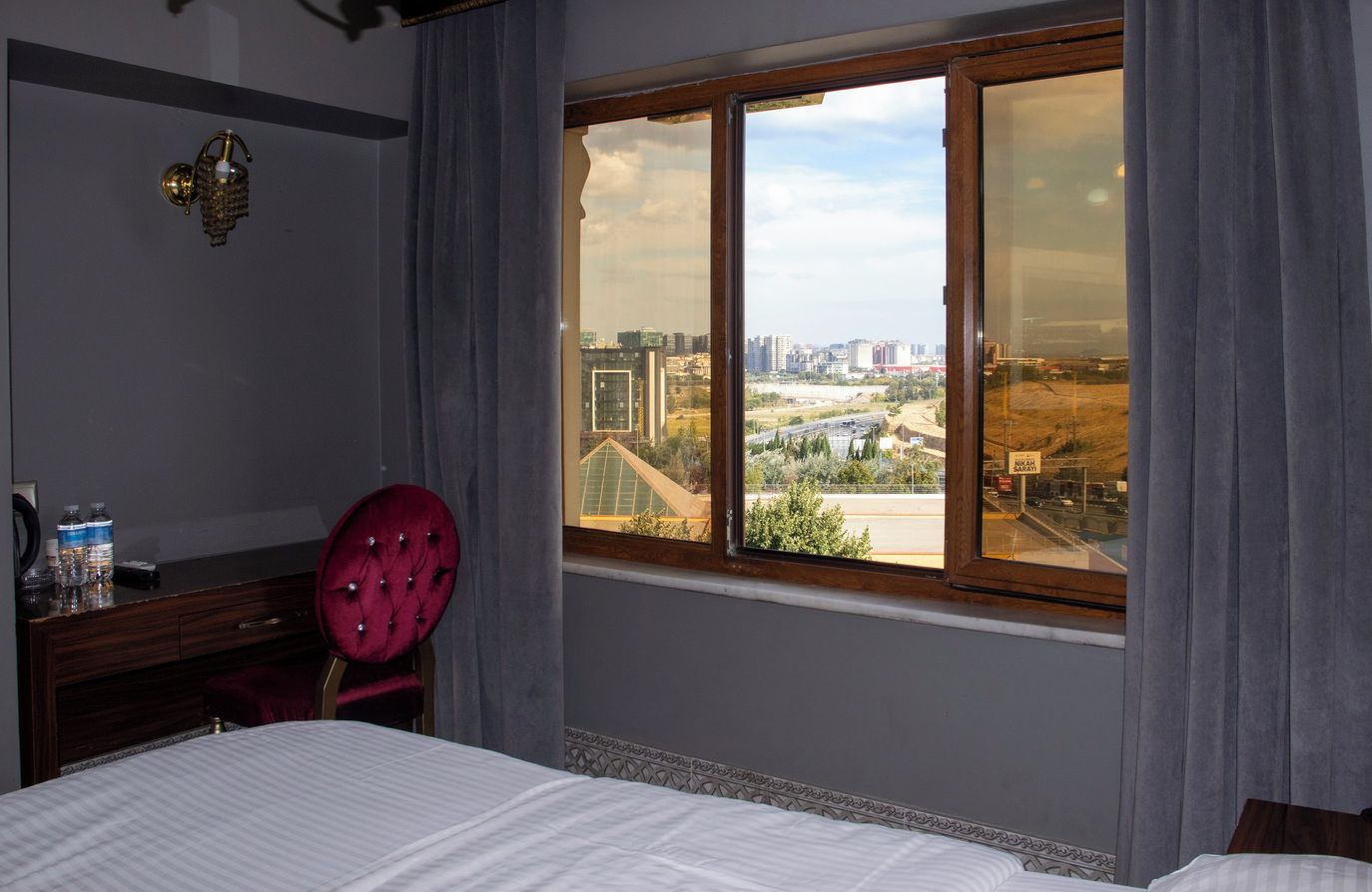 Quarto de hotel com uma janela grande, uma cama, uma escrivaninha com cadeira e uma luminária de parede decorativa.