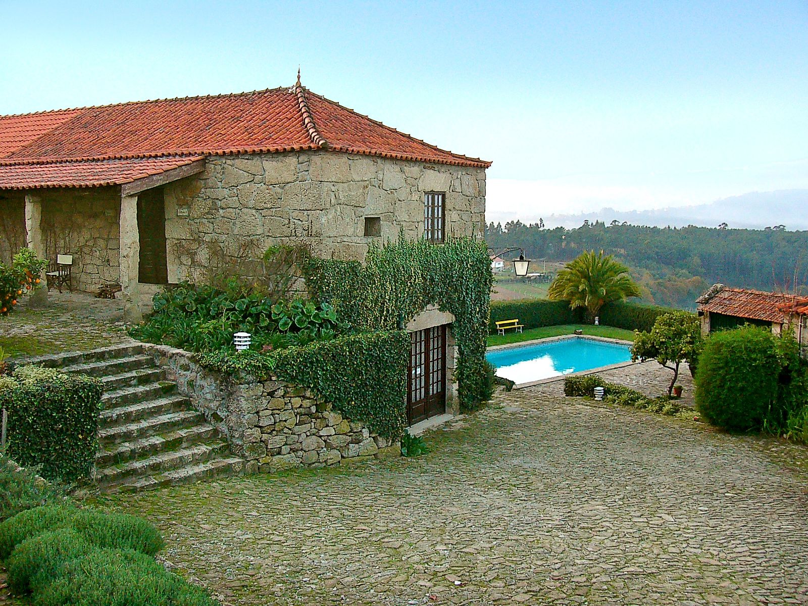 Um edifício de pedra com telhado em telhas, uma piscina e uma escadaria de pedra que leva à entrada.