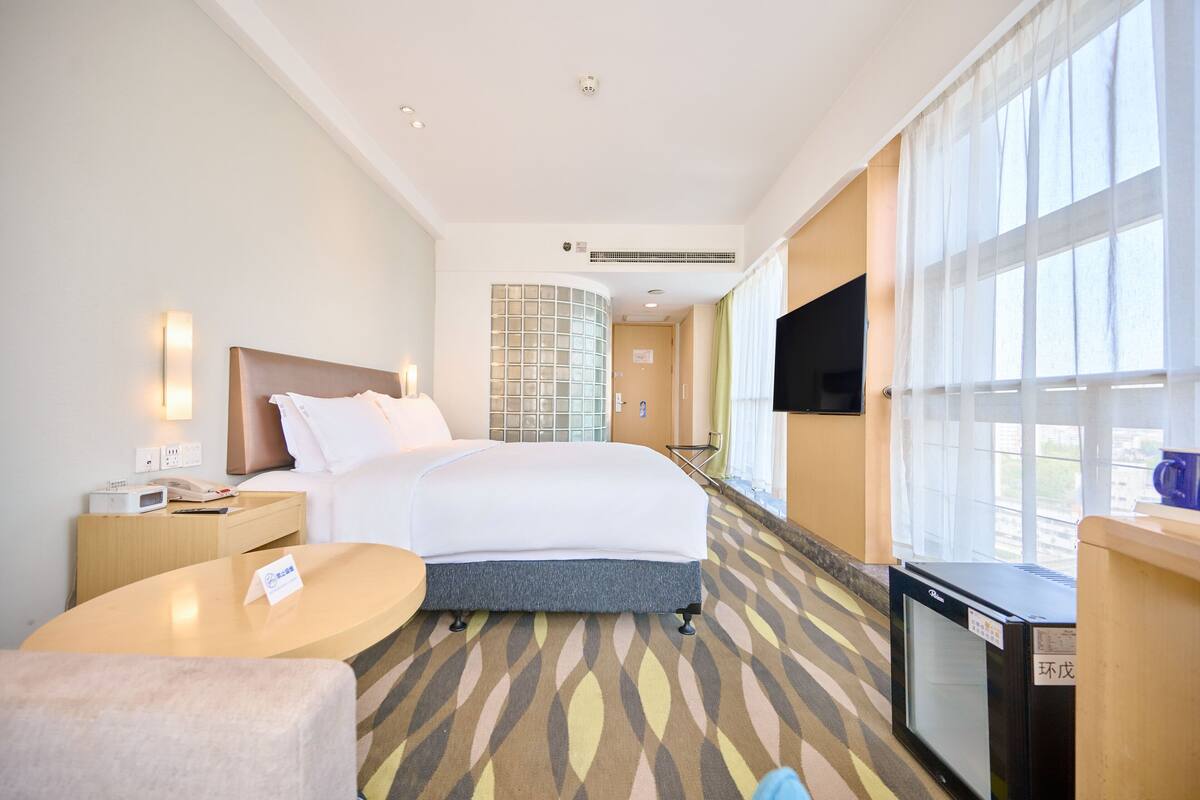 Quarto de hotel com cama, mesa de cabeceira, abajur, televisão e pequena escrivaninha com computador.