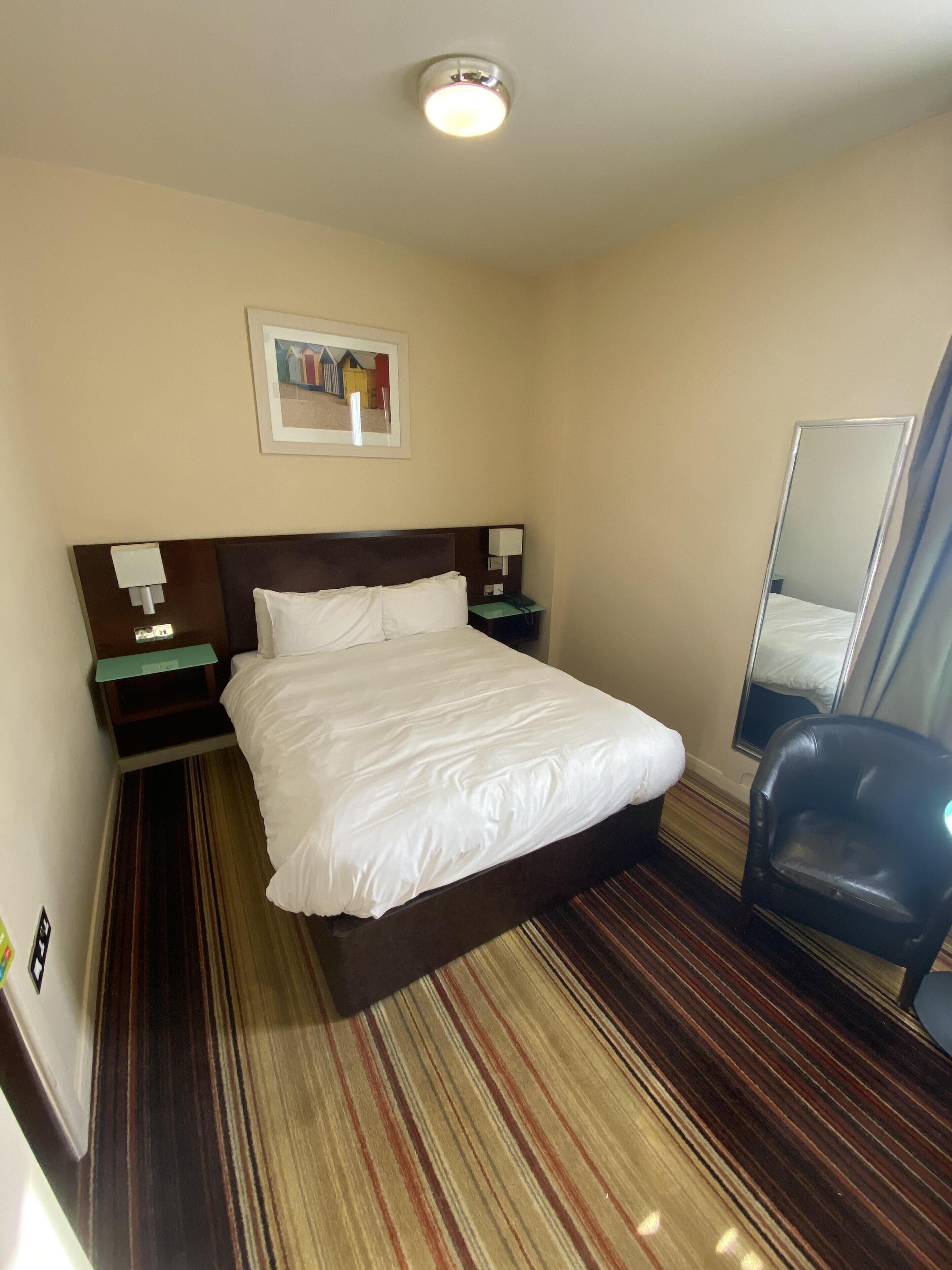 Quarto de hotel com uma cama, mesa de cabeceira, abajur, quadro na parede, espelho e tapete listrado.