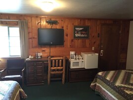 Quarto com parede revestida em madeira, televisão fixada acima de uma escrivaninha, uma cadeira, uma cama e um refrigerador.
