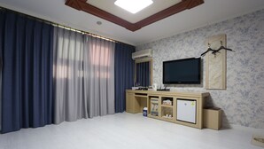 Quarto com uma janela grande, cortinas, TV de tela plana, escrivaninha de madeira e uma pequena geladeira.
