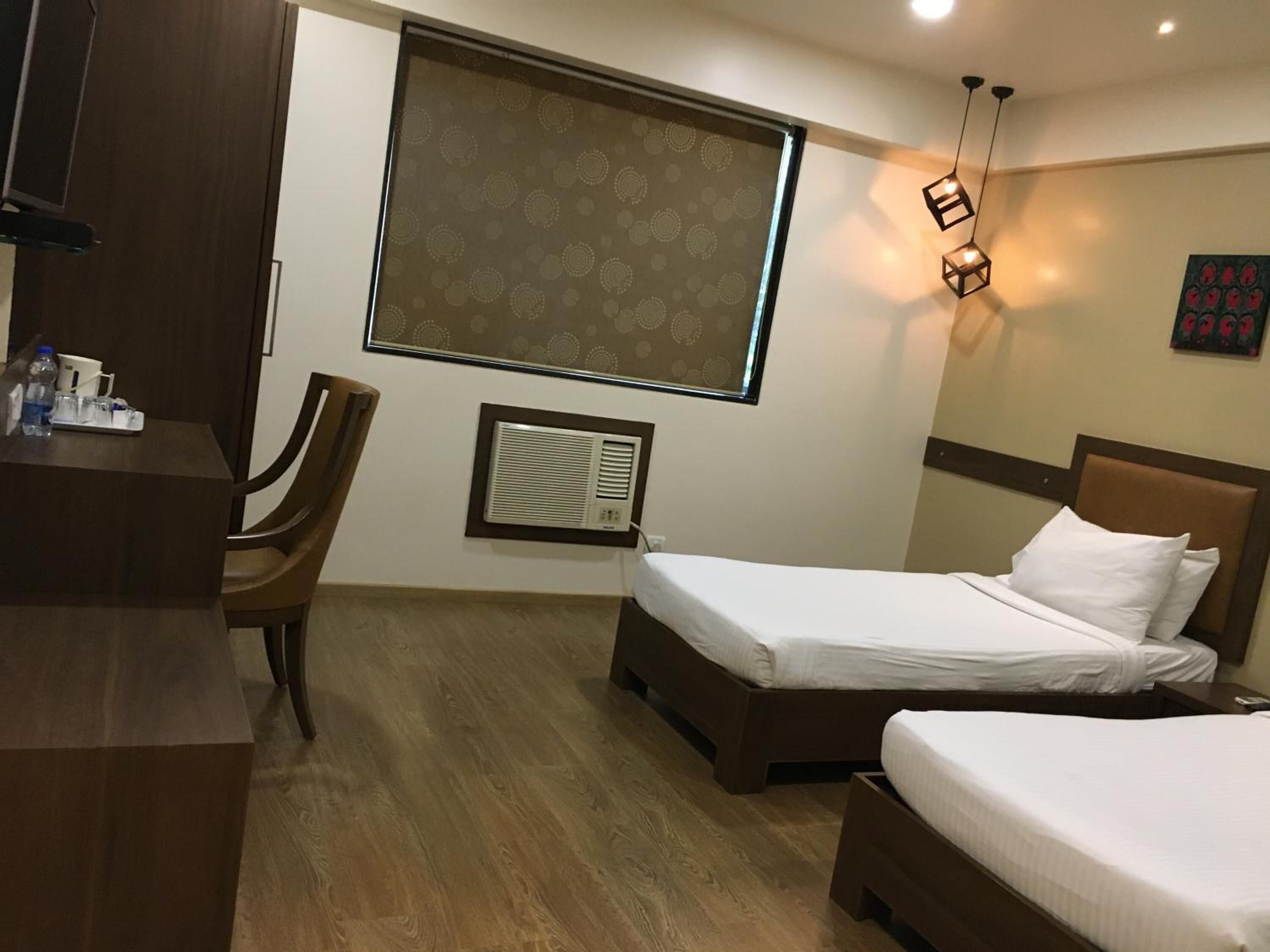 Quarto de hotel com duas camas, uma escrivaninha e uma janela com persiana estampada.