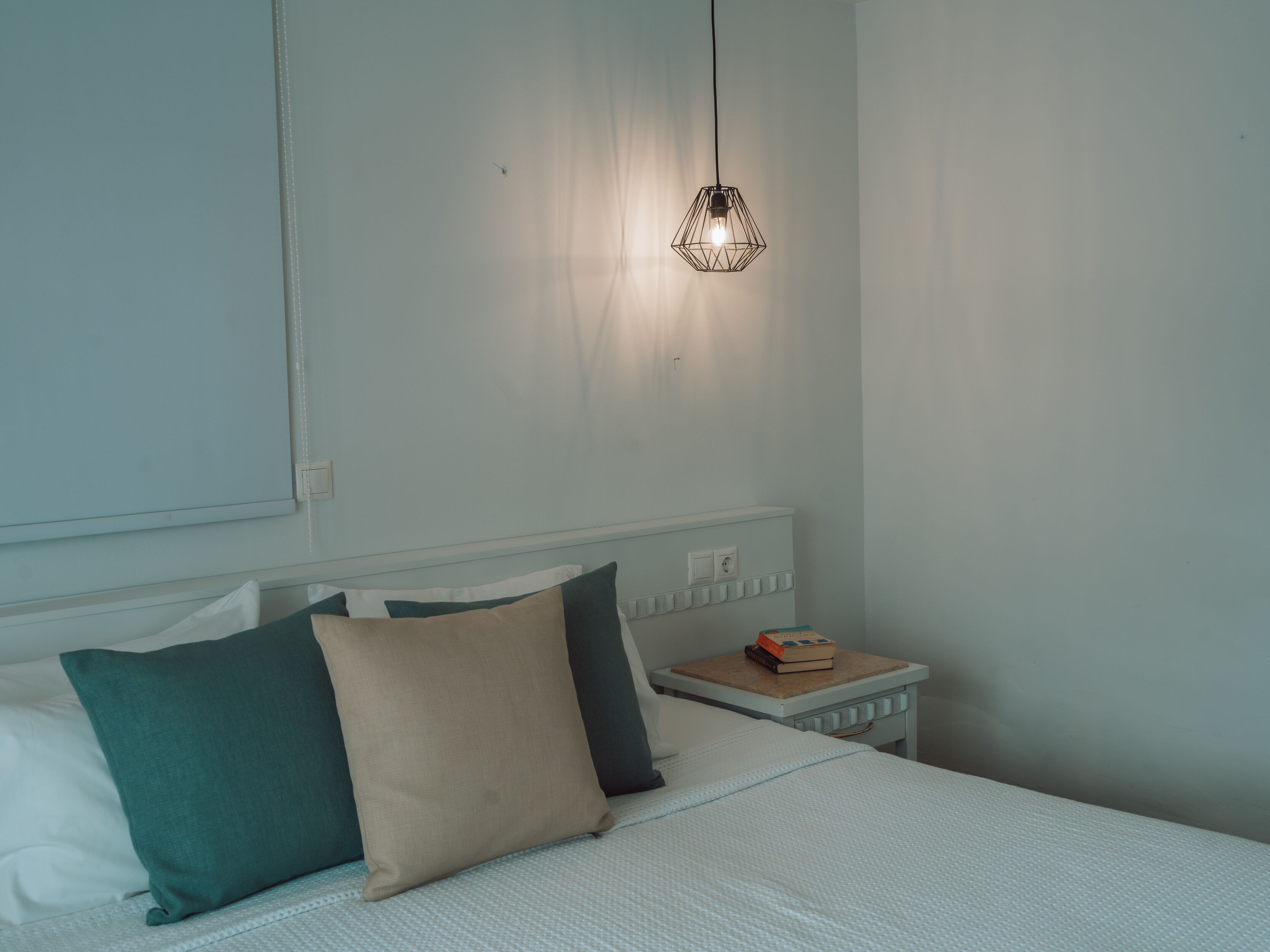 Uma cama bem arrumada com travesseiros, uma mesa de cabeceira com livros e uma luminária pendente.