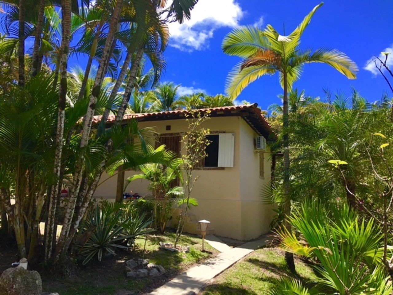 Um pequeno edifício térreo com telhado de telhas, rodeado por vegetação tropical e palmeiras.