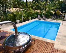 Piscina com grade metálica, rodeada por um deck de madeira e espreguiçadeiras, com vegetação exuberante ao fundo.