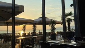 Restaurante com grandes janelas voltadas para um pôr do sol, com mesas ao ar livre e vasos de plantas.