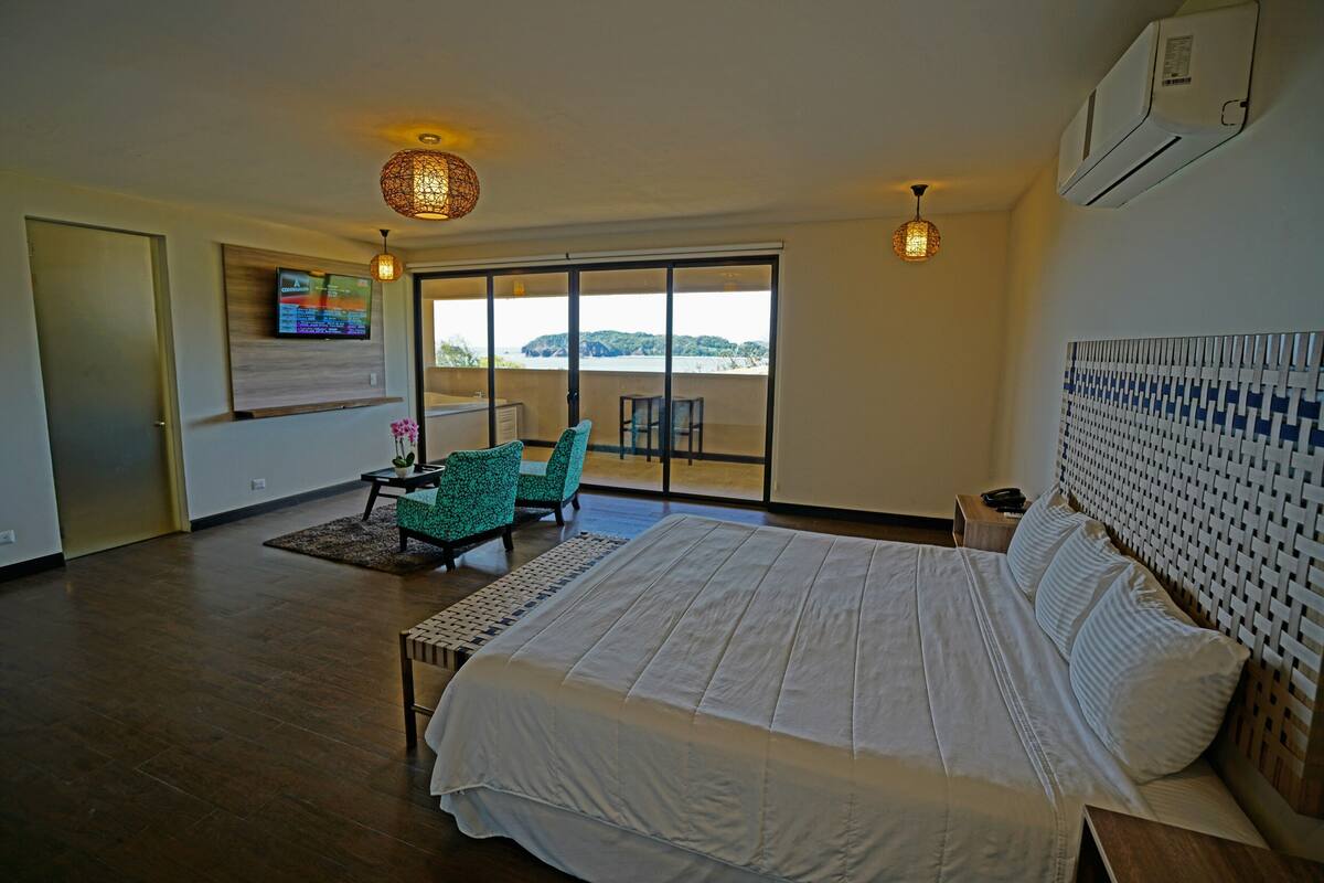 Quarto com uma cama grande, uma cadeira, uma televisão e vista para o exterior.