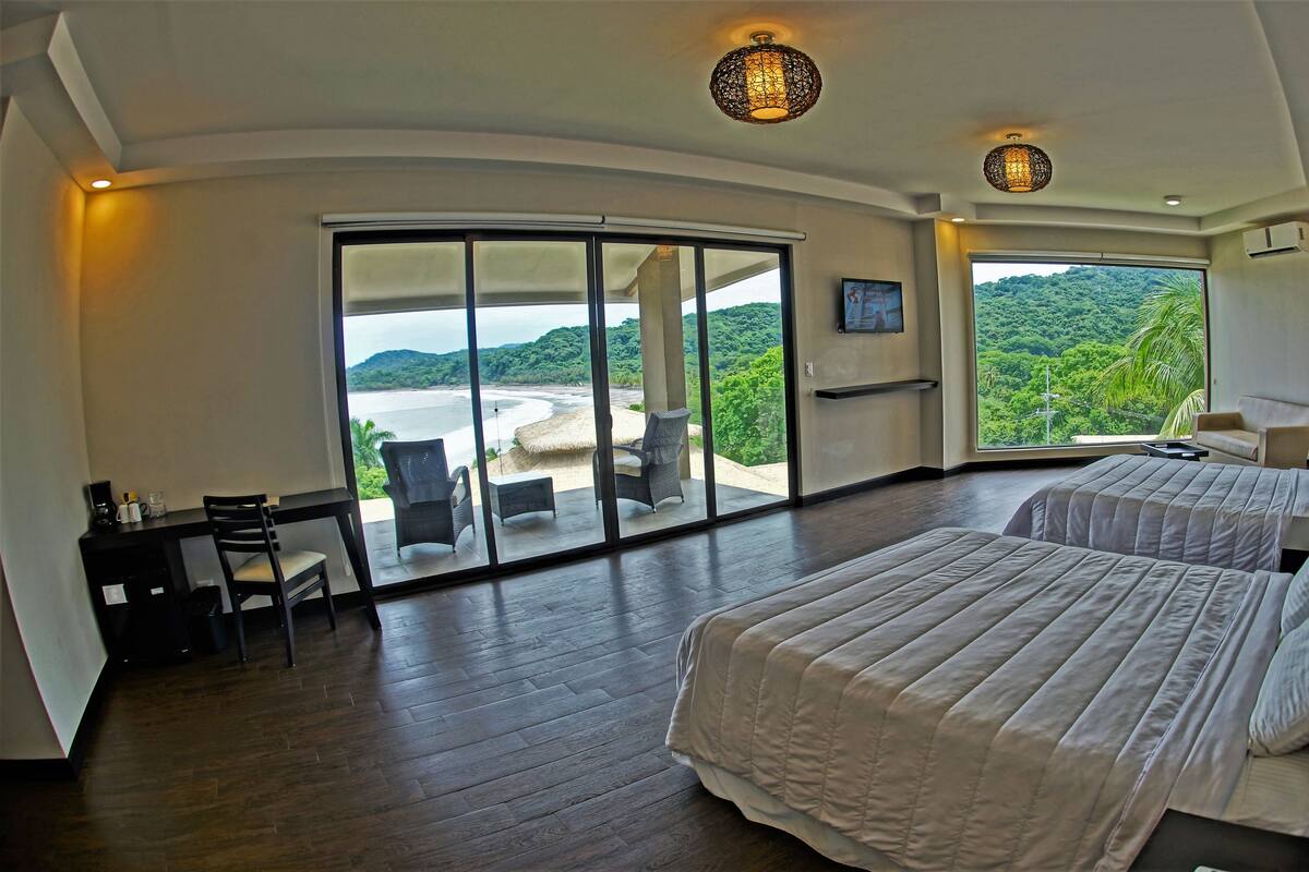 Quarto com duas camas, uma escrivaninha e uma cadeira, com uma grande porta de vidro deslizante que dá para uma varanda com vista para a praia e montanhas.