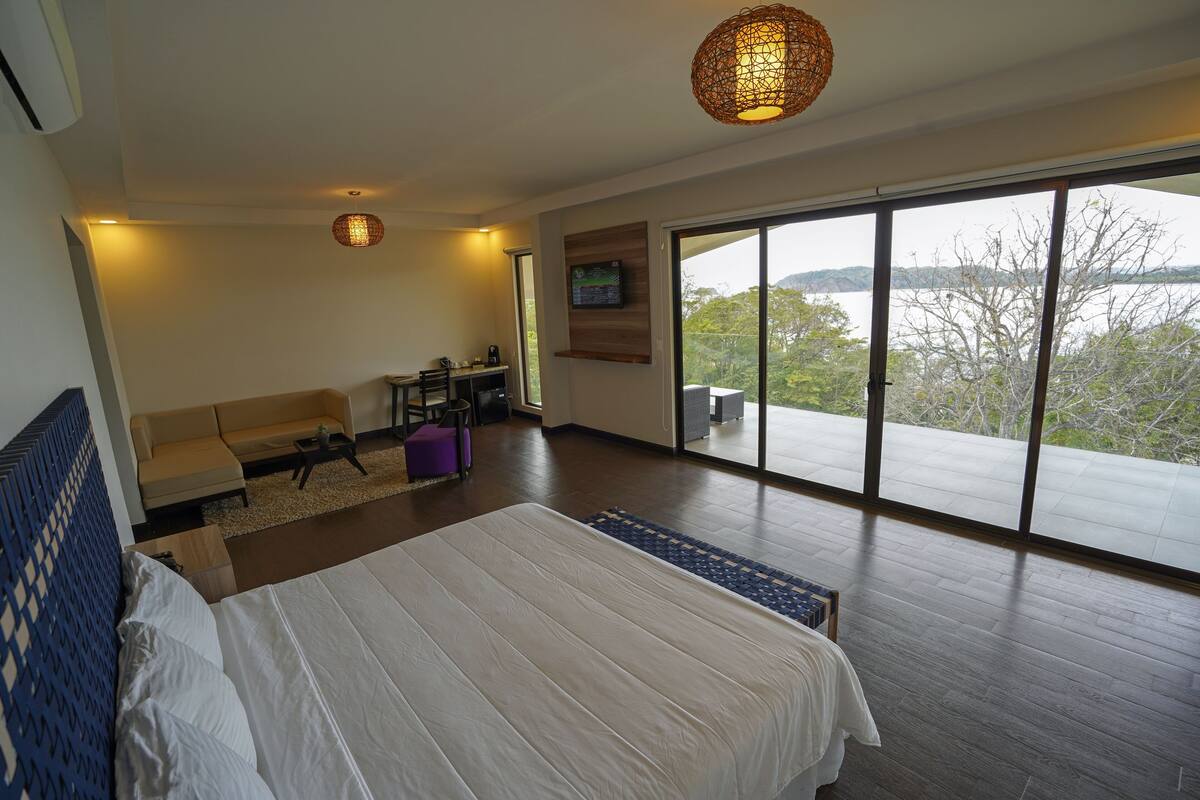 Quarto com uma cama grande, um sofá, uma TV fixada na parede e vista para o exterior através de uma porta de vidro deslizante.