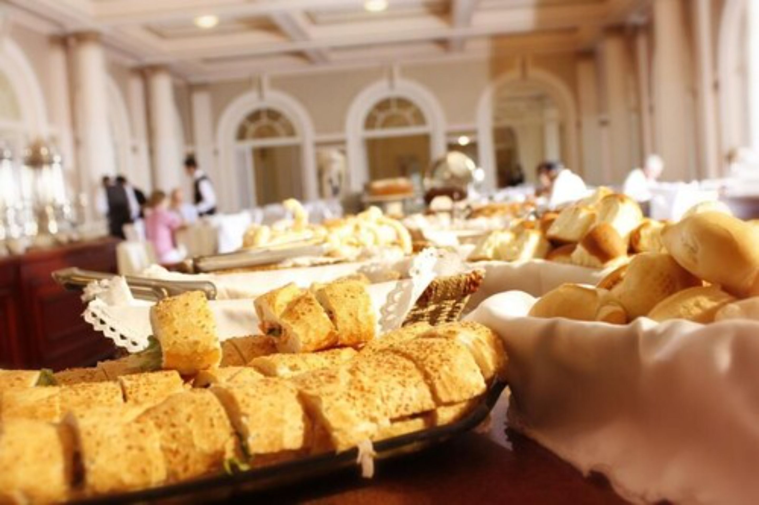 Buffet com diversos alimentos, incluindo pão e doces, em um salão com janelas em arco e decoração clássica.