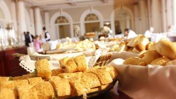 Buffet com diversos alimentos, incluindo pão e doces, em um salão com janelas em arco e decoração clássica.