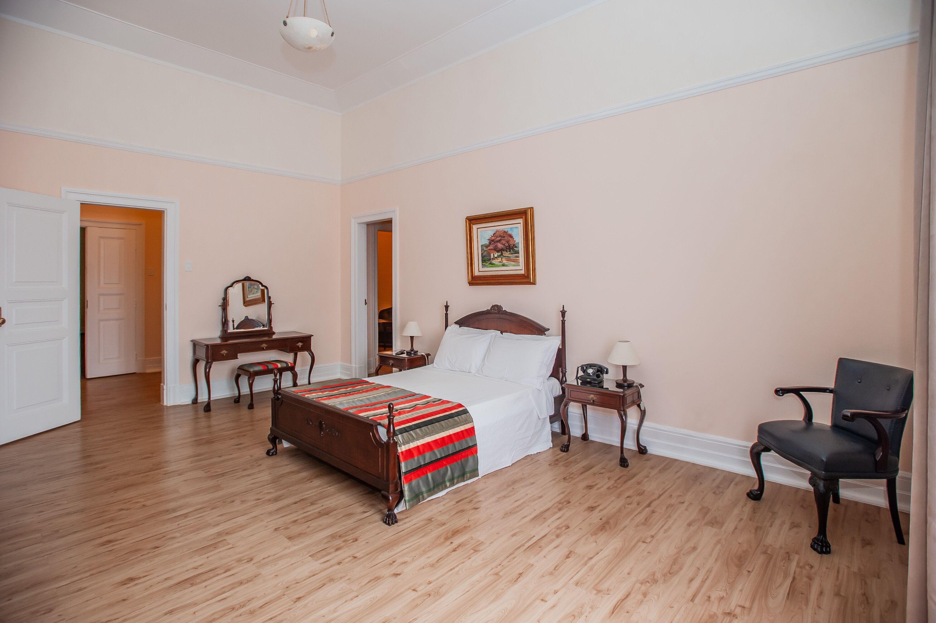 Quarto com cama de madeira, uma cadeira, mesa de cabeceira, cômoda e um quadro na parede.