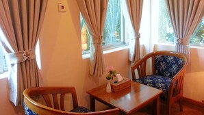 Quarto com duas cadeiras de madeira, uma mesa pequena, cortinas bege e janela com vista para o verde.