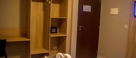 Quarto de hotel com cama, guarda-roupa de madeira, secretária e porta com placa.