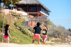 Jogo de voleibol de praia com jogadores em primeiro plano e um edifício de telhado de palha ao fundo.