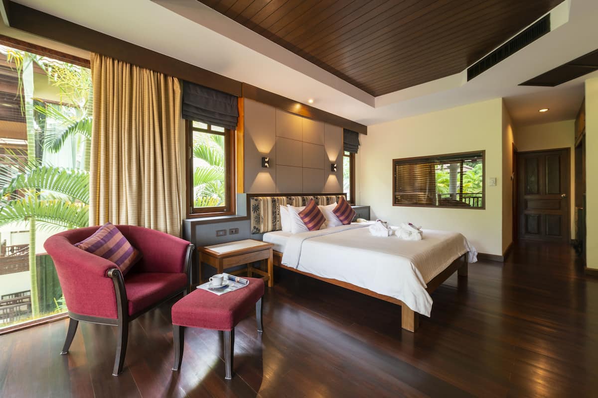 Um quarto espaçoso com uma cama grande, uma poltrona vermelha, uma mesa de cabeceira de madeira e vista para o exterior.