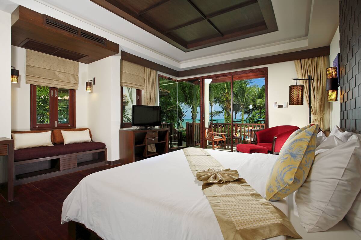 Quarto de hotel com uma cama grande, uma escrivaninha com computador, uma cadeira vermelha e vista para palmeiras e oceano.