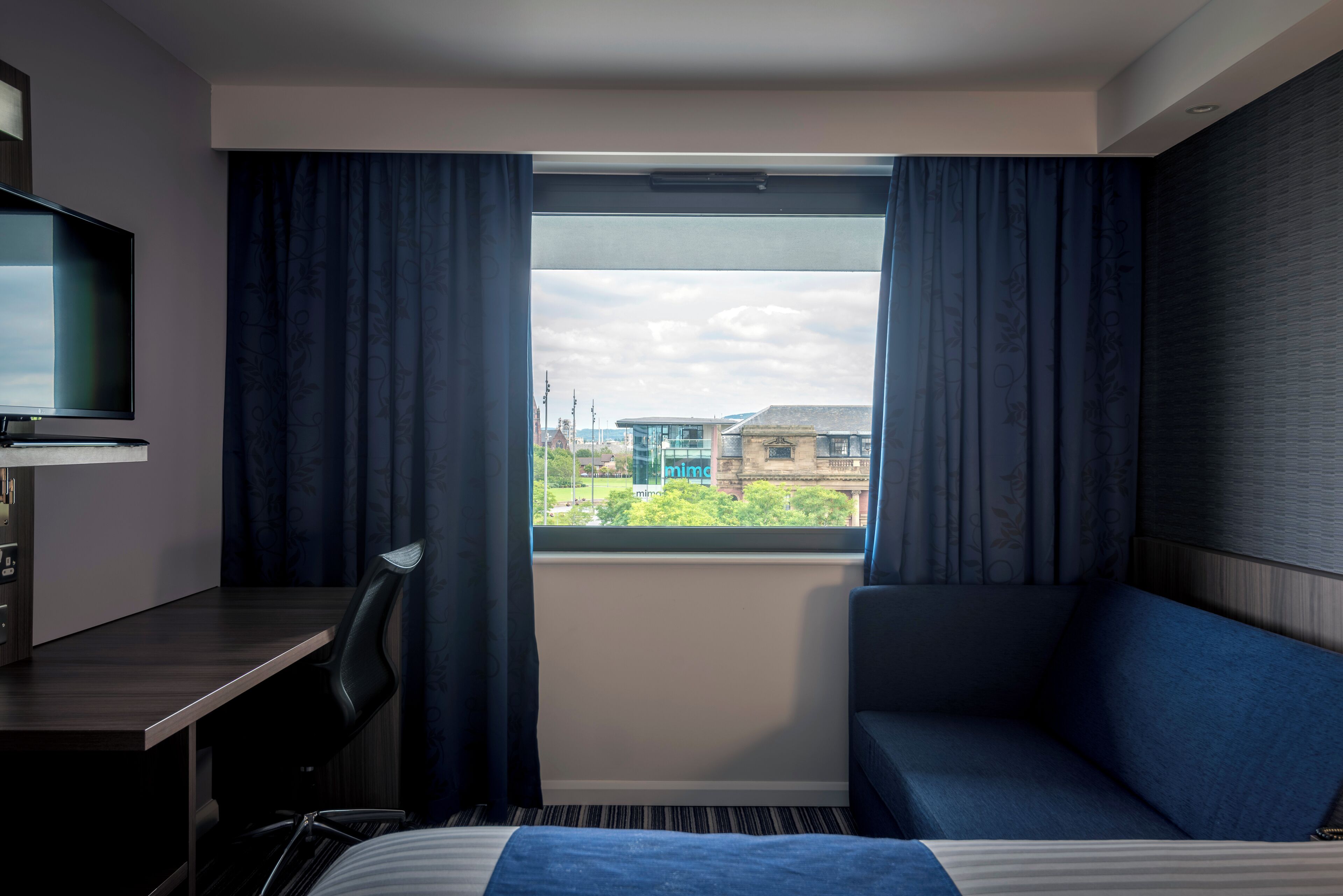 Quarto de hotel com cama, escrivaninha, cadeira, televisão e janela com cortinas.