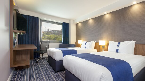 Quarto de hotel com duas camas, uma escrivaninha, uma cadeira, uma televisão e uma janela com vista para a cidade.