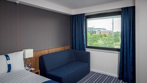 Quarto de hotel com cama, sofá, mesa de cabeceira com abajur e janela com cortinas.