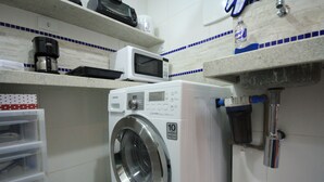 Uma área de lavanderia compacta com máquina de lavar de abertura frontal, micro-ondas e cafeteira sobre a bancada.