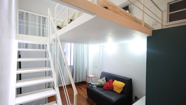 Um quarto com uma cama de beliche, um sofá preto com almofadas coloridas e uma pequena mesa com livros.