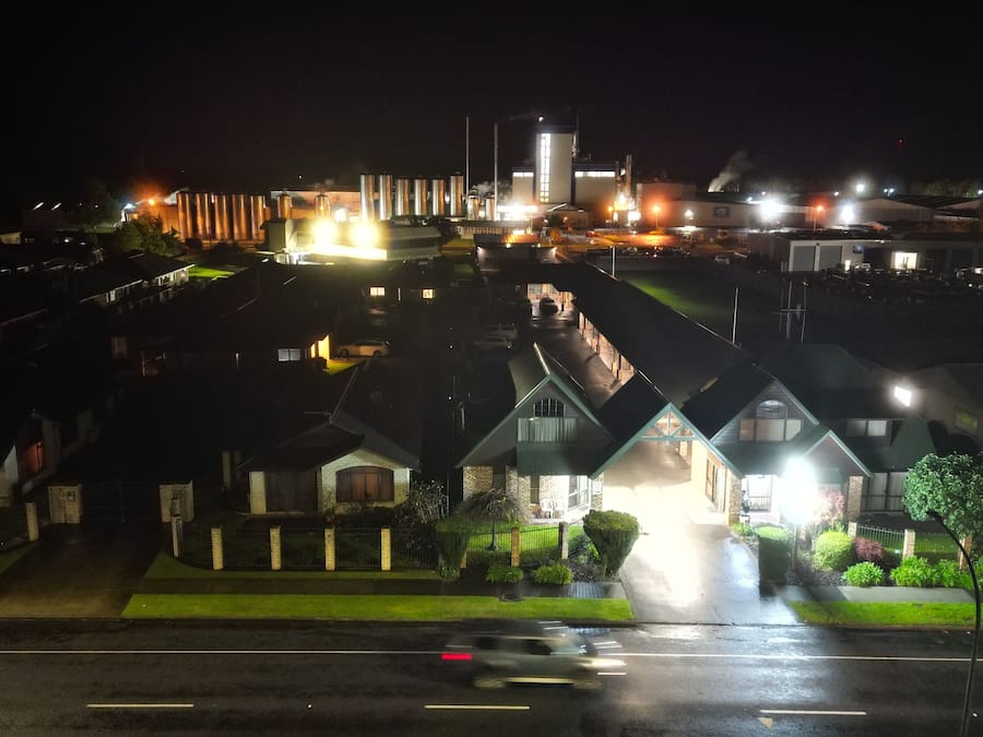 Vista noturna de uma área residencial com casas e uma grande instalação industrial ao fundo.