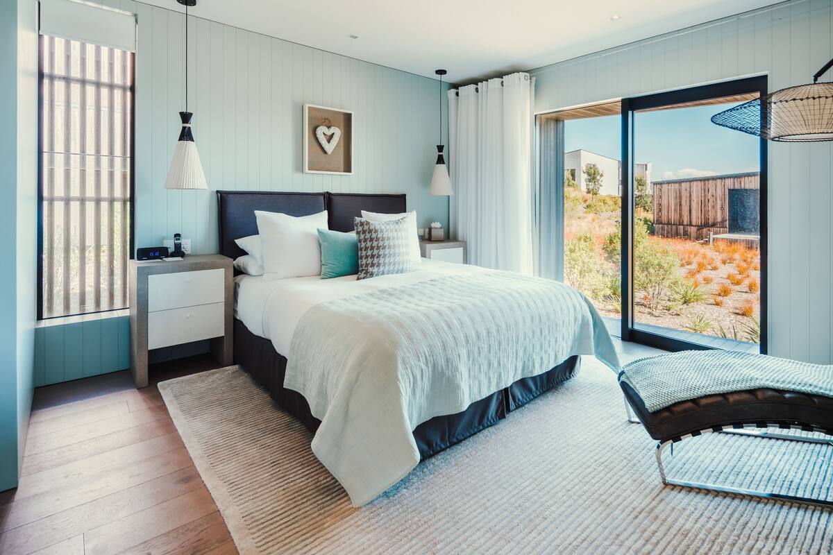 Quarto com cama, mesas de cabeceira, mesa de apoio, janela com cortinas e vista para o exterior.