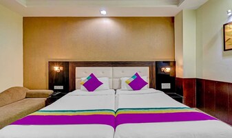 Uma cama de casal com um edredom colorido listrado e duas almofadas, uma mesinha de cabeceira com abajur, e um sofá em um quarto de hotel.